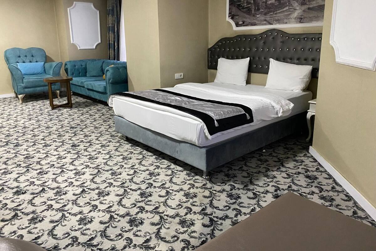 Quarto de hotel com uma cama grande, uma poltrona azul, uma mesinha de apoio e um tapete estampado.