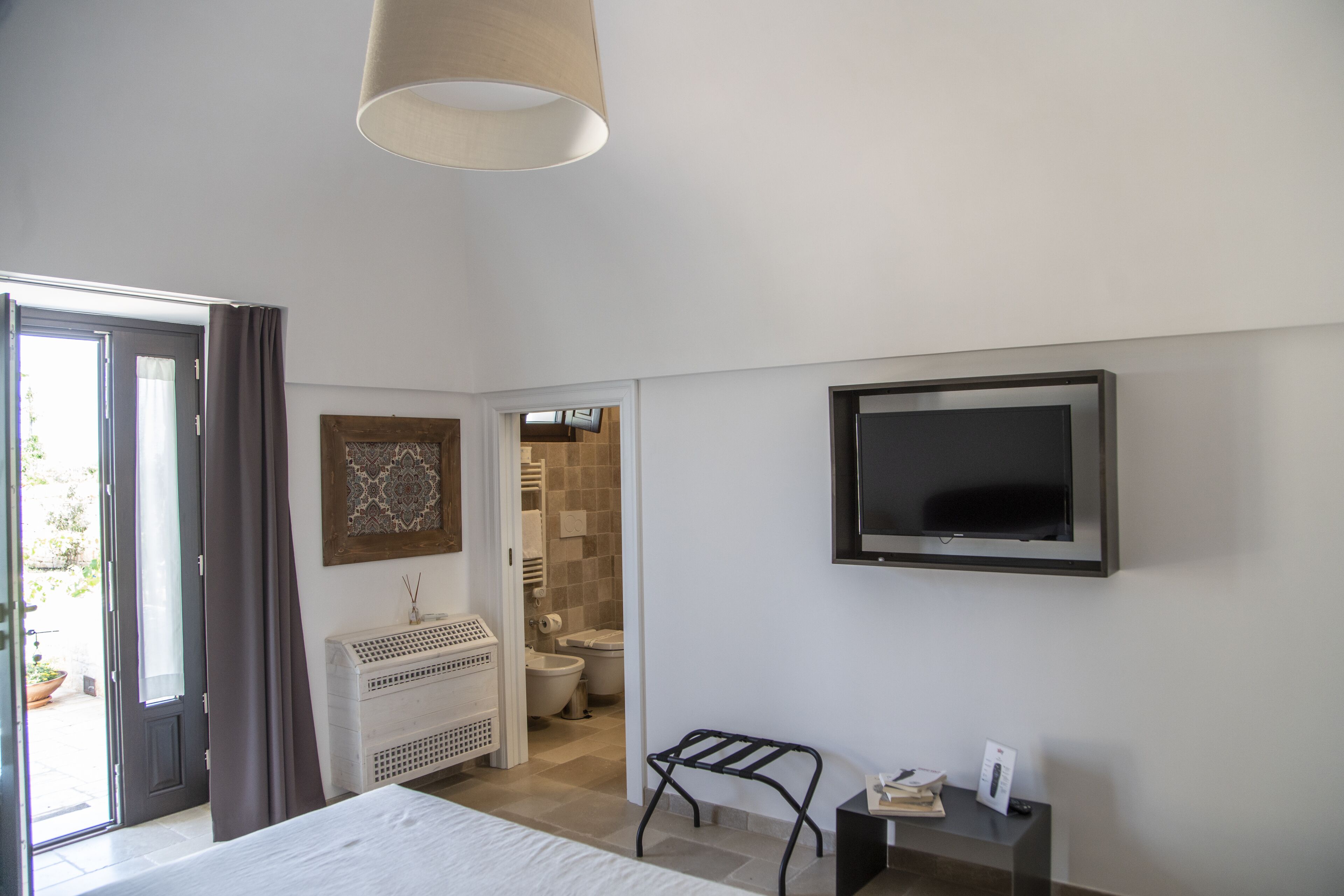Quarto de hotel com uma cama, televisão na parede, uma mesa pequena com um controle remoto e uma porta que dá para um banheiro.