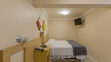 Quarto com cama, mesa de cabeceira, luminária e televisão fixada na parede.