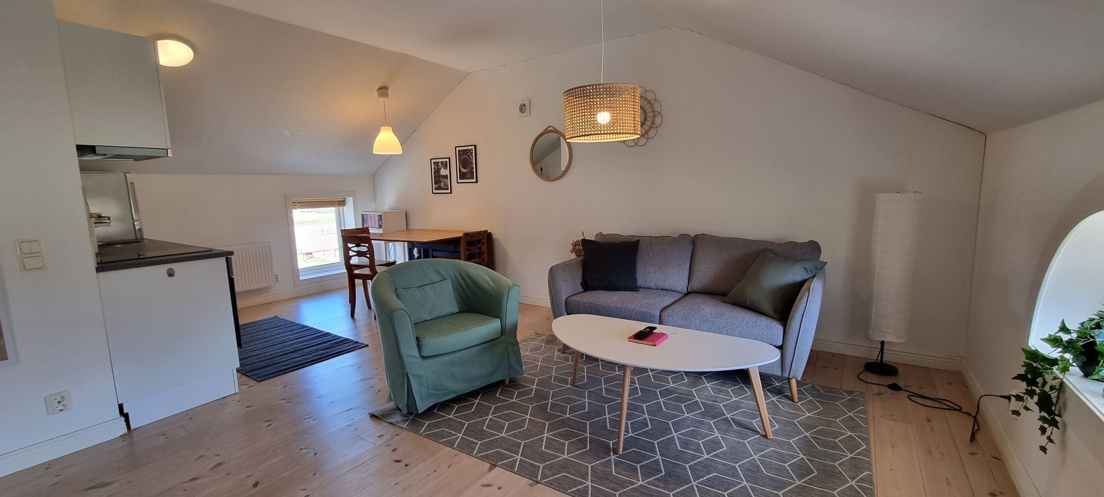 Uma sala de estar moderna com um sofá cinzento, uma poltrona verde e uma mesa de centro branca. Há uma área de cozinha com fogão e uma mesa de jantar com cadeiras.