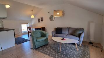 Uma sala de estar moderna com um sofá cinzento, uma poltrona verde e uma mesa de centro branca. Há uma área de cozinha com fogão e uma mesa de jantar com cadeiras.
