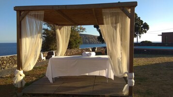 Um gazebo de madeira com uma mesa e cadeiras brancas, adornado com cortinas leves, com vista para uma parede de pedra e uma piscina.
