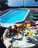 Disposição para o café da manhã ao lado de uma piscina, com toalha de mesa, alimentos para o café e vista para a piscina.