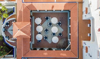 Vista aérea de um pátio com piso de azulejos, cercado por edifícios com telhados de terracota. Há várias mesas redondas com cadeiras e guarda-sóis brancos, e uma área central com um grande guarda-sol branco.