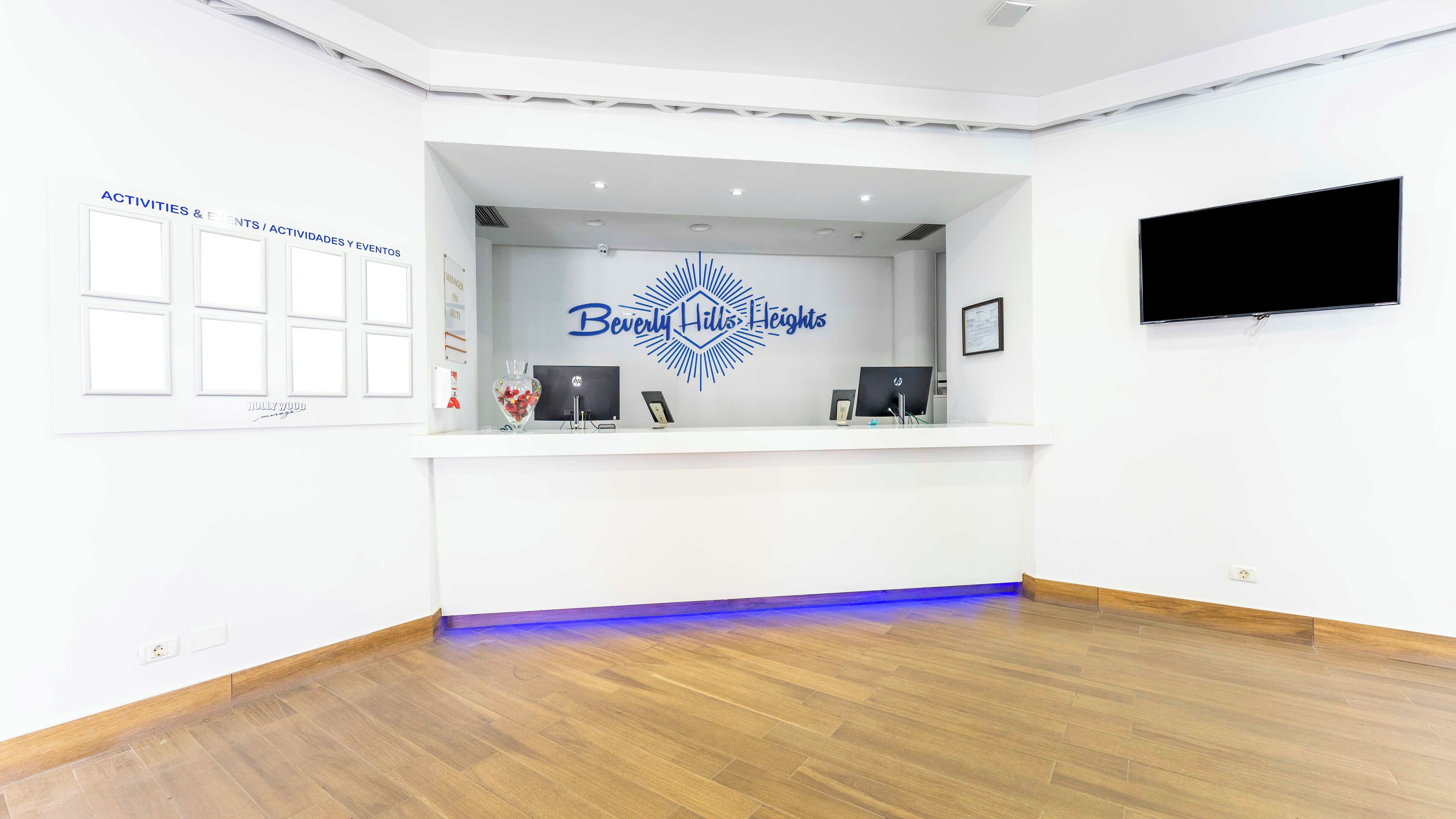 Balcão de recepção com o logotipo Beverly Hills Heights, dois monitores HP e uma TV de tela plana.