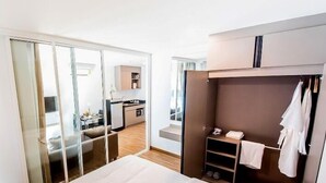 Quarto de hotel moderno com cama, guarda-roupa, pequeno espaço de estar e cozinha compacta.