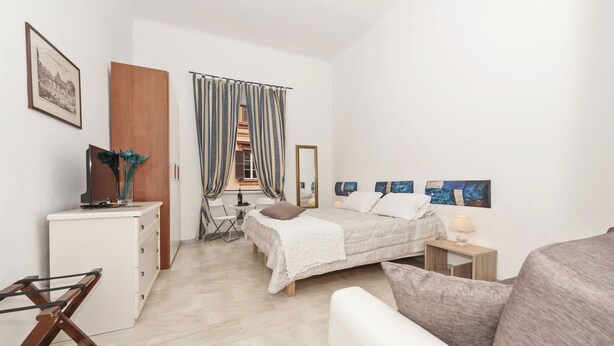 Quarto com cama, mesas de cabeceira, escrivaninha, cadeira e janela com cortinas.