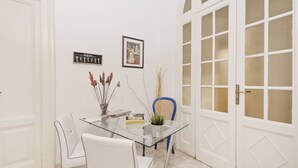 Uma área de refeições com mesa de vidro, cadeiras brancas, uma cadeira azul, um vaso de planta, um quadro e um aviso com chaves.