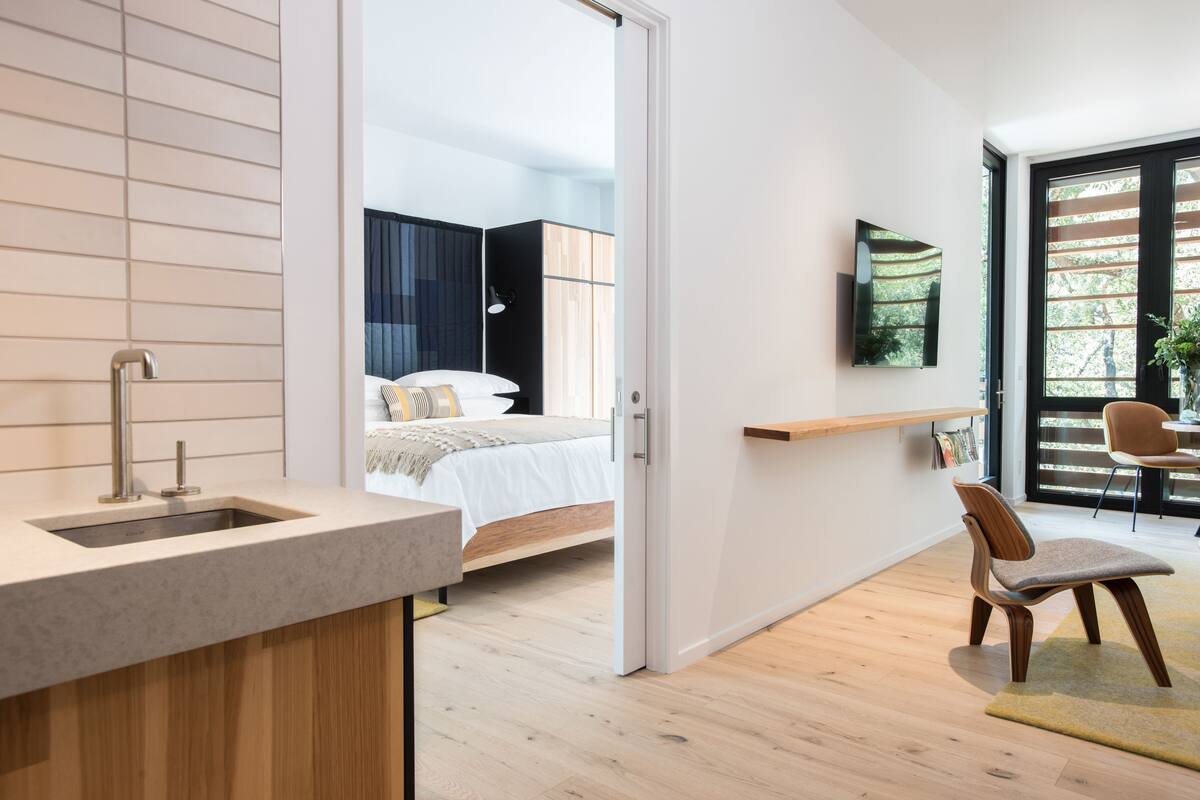 Quarto de hotel moderno com piso de madeira, uma cama grande, escrivaninha embutida, uma cadeira e uma janela ampla.