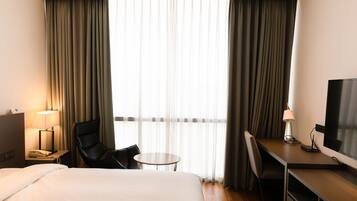Quarto de hotel com cama, escrivaninha, cadeira, televisão e janela com cortinas.