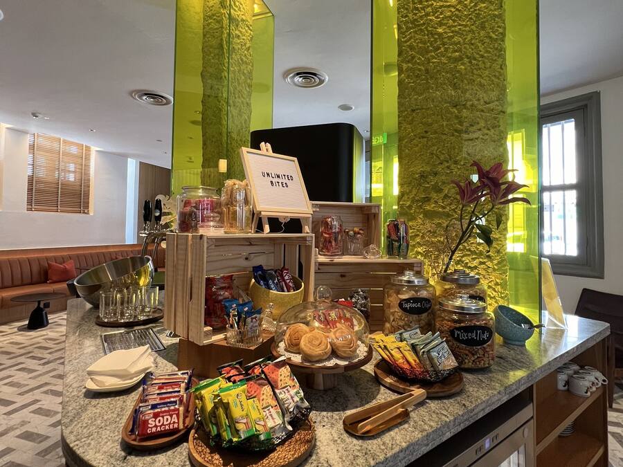 Uma área de buffet com diversos alimentos, incluindo potes de mix de frutos secos e petiscos, uma cesta de frutas, e uma placa com a inscrição 'Petiscos Ilimitados'.