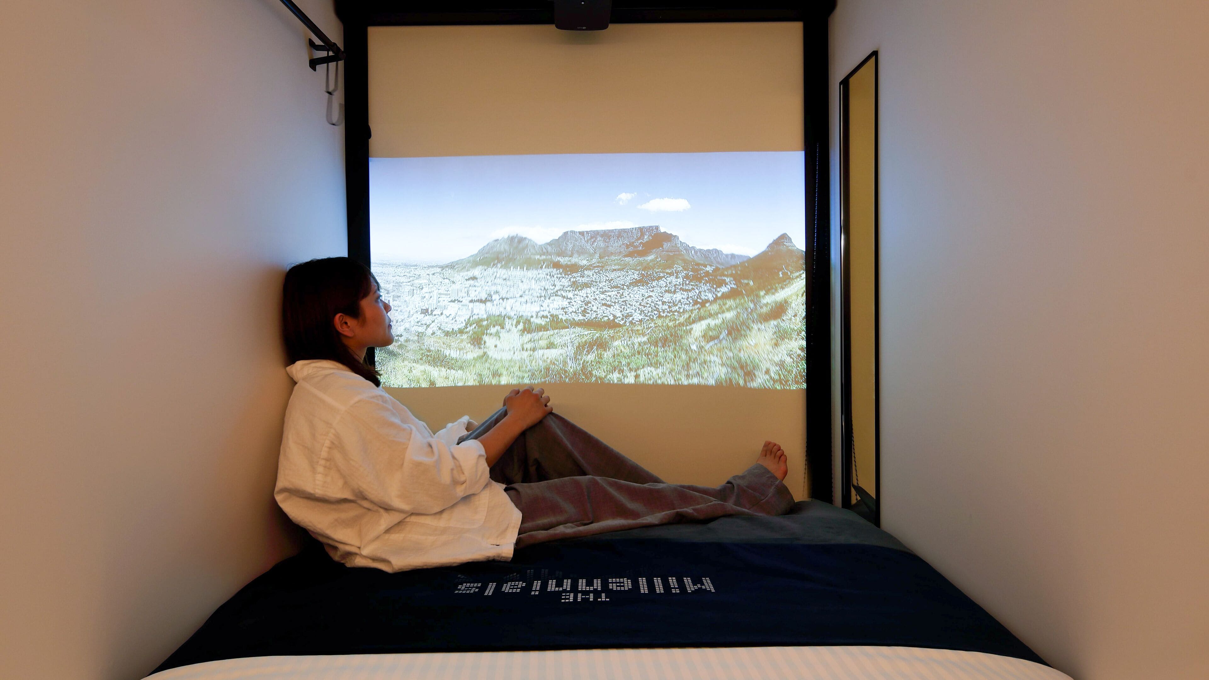 Uma pessoa sentada em uma cama, com projeção de uma paisagem montanhosa na parede.