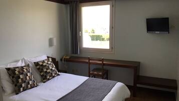 Quarto de hotel com cama, escrivaninha, cadeira, televisão e janela com cortinas.