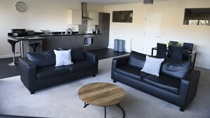 Uma sala de estar moderna com dois sofás de couro preto, uma mesa de centro de madeira e uma cozinha ao fundo.