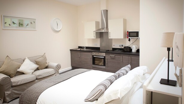 Quarto de hotel moderno com cama, sofá, cozinha compacta e um relógio na parede.