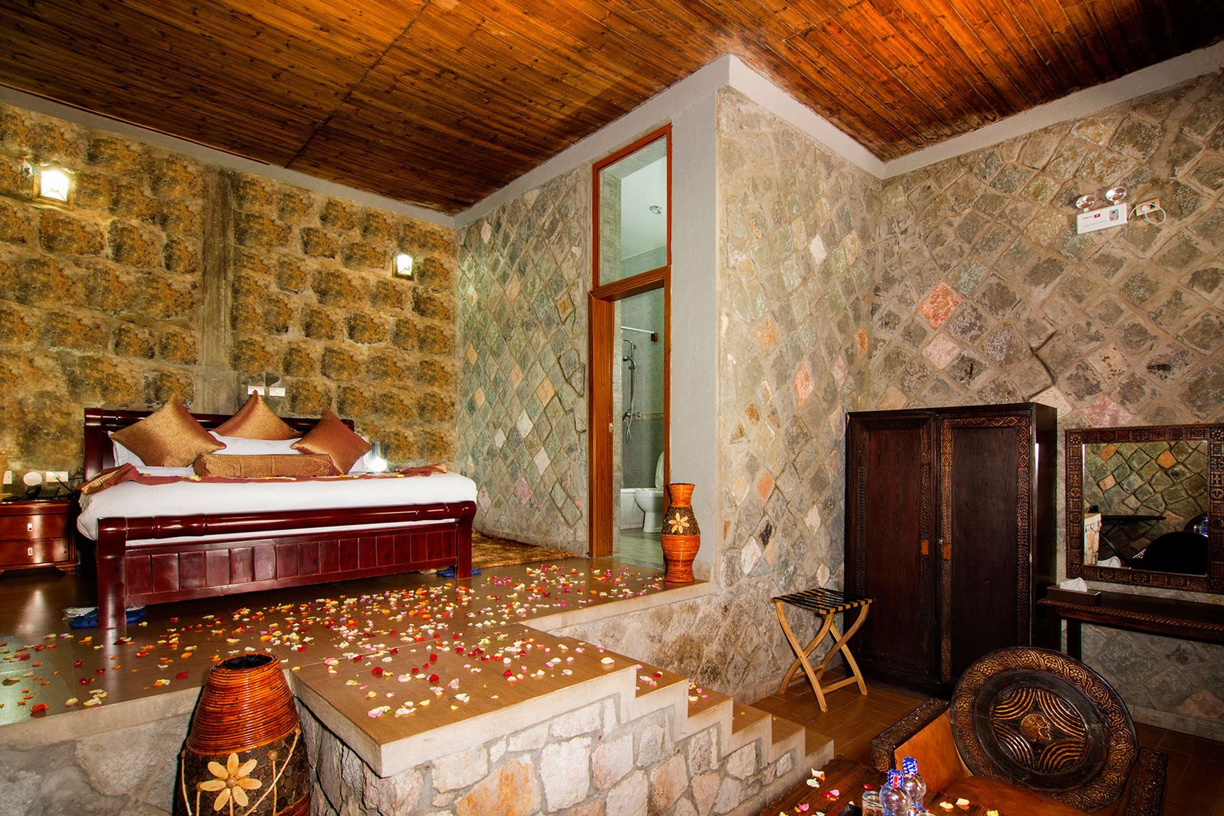 Quarto com parede de pedra, teto de madeira, cama com almofadas, armário de madeira e pequena mesa com um vaso.