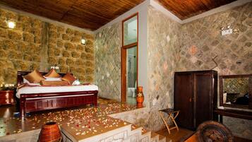 Quarto com parede de pedra, teto de madeira, cama com almofadas, armário de madeira e pequena mesa com um vaso.