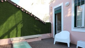 Área de pátio com uma pequena piscina, uma cadeira branca e uma parede rosa com um painel de grama sintética verde.