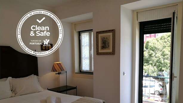 Quarto de hotel com uma cama, uma mesa de cabeceira, uma luminária, uma janela e uma porta que dá para uma varanda com vista para carros estacionados e vegetação.