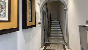 Um corredor com escada que leva a outro espaço. Quadros emoldurados decoram as paredes e há uma porta com maçaneta.
