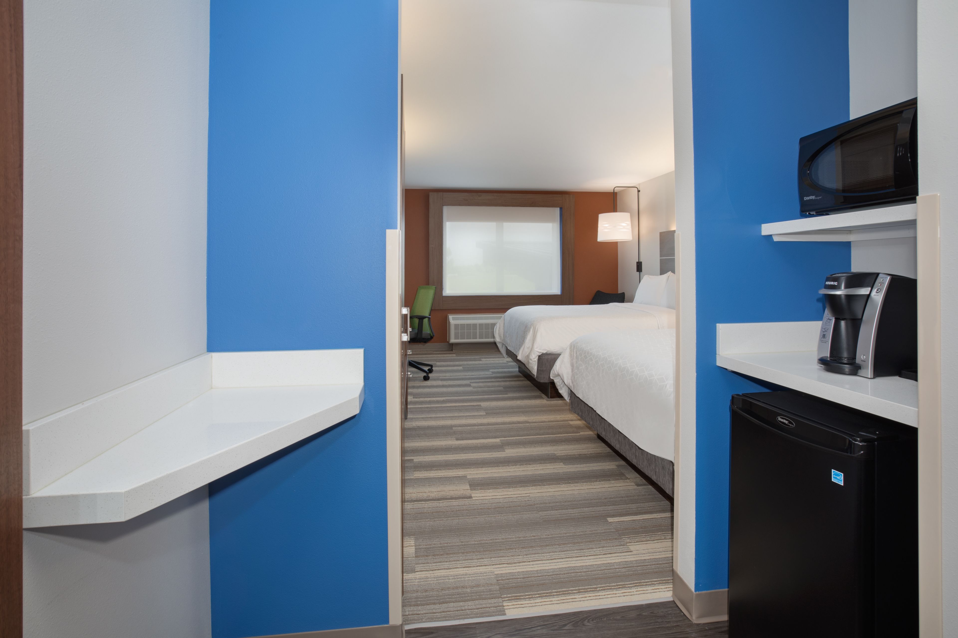 Quarto de hotel com cama, escrivaninha com micro-ondas, máquina de café e pequena geladeira.
