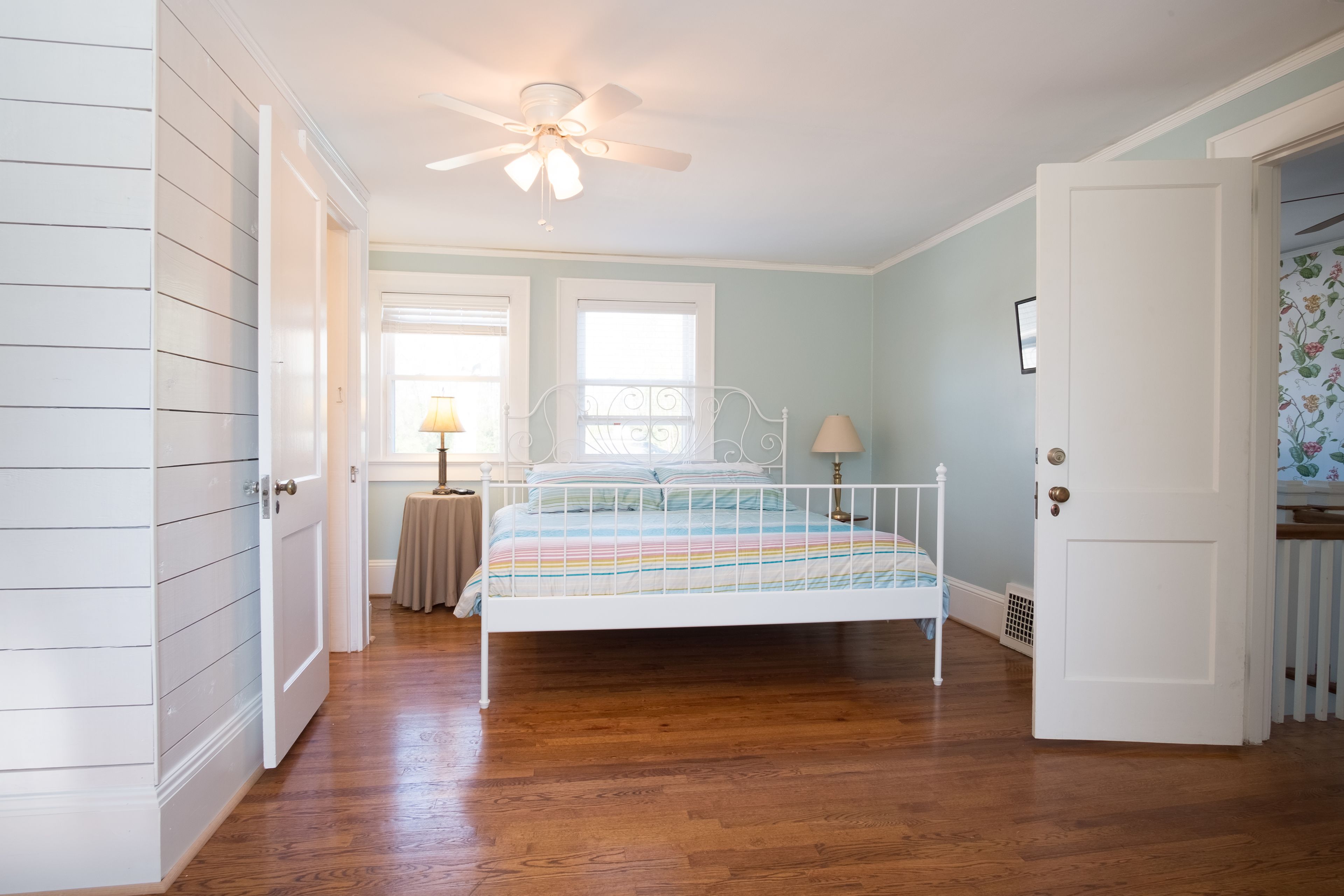 Quarto com estrutura de cama em metal branco, ventoinha no teto, duas janelas com molduras brancas, uma porta e um radiador.