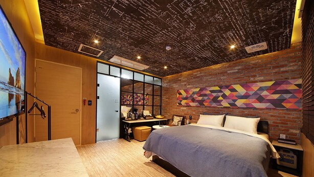 Quarto de hotel moderno com uma cama grande, uma mesa e um espelho. O quarto possui uma parede de tijolos e um teto com padrão geométrico.