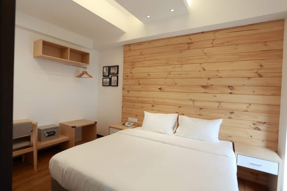 Quarto de hotel moderno com cabeceira de madeira, cama com roupa branca, escrivaninha com cadeira, uma pequena prateleira e mesa de cabeceira com telefone.
