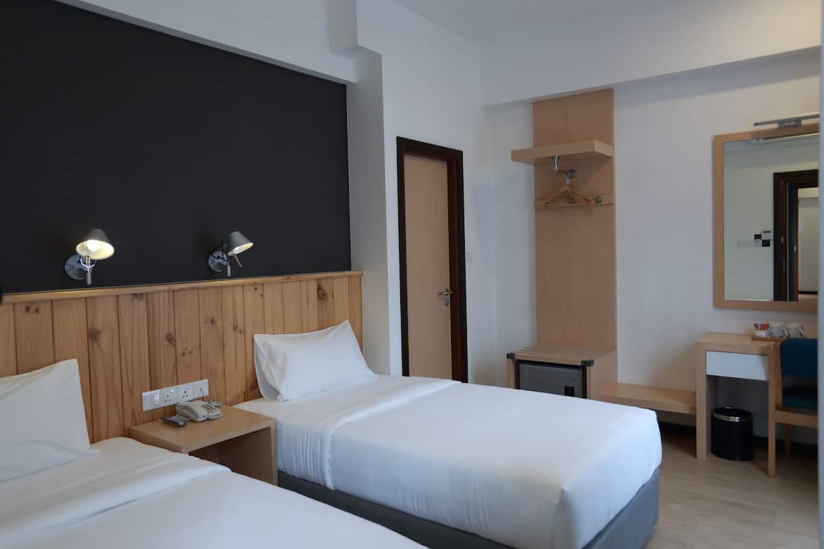 Quarto de hotel com duas camas, cabeceira de madeira, mesas de cabeceira com luminárias, um espelho, uma escrivaninha com cadeira e um cesto de lixo pequeno.