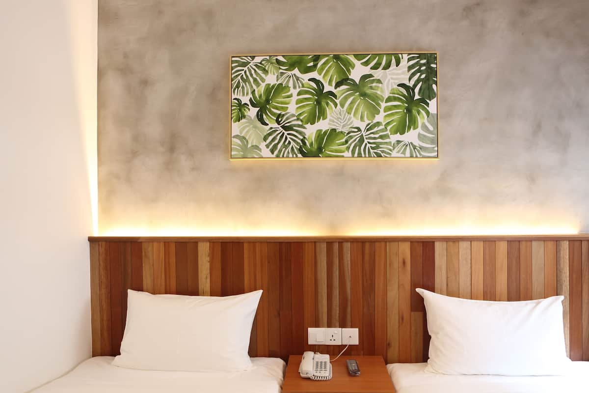 Quarto de hotel com cabeceira de madeira, duas almofadas brancas, mesa de cabeceira com telefone e controle remoto, e uma obra de arte na parede com padrão de folhagem tropical.