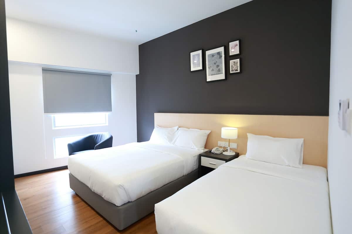 Quarto de hotel com duas camas, uma janela grande com cortinas, parede escura com quadros e uma mesa de cabeceira com abajur.