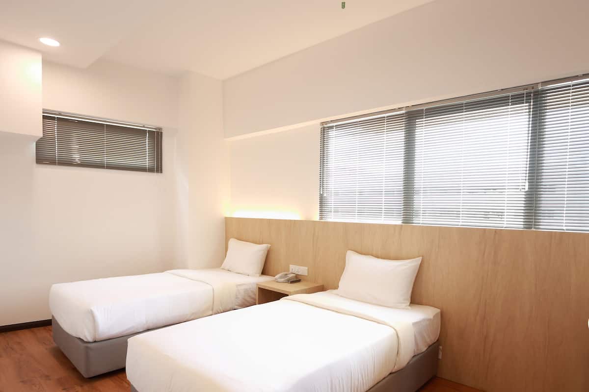 Quarto de hotel com duas camas de solteiro, cabeceira de madeira, mesa de cabeceira com telefone e janela com persianas.