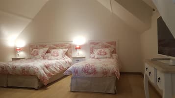 Quarto com duas camas, cada uma com uma mesa de cabeceira e uma luminária. Há um penteado com espelho e uma televisão em cima. O quarto tem teto inclinado e piso de madeira.