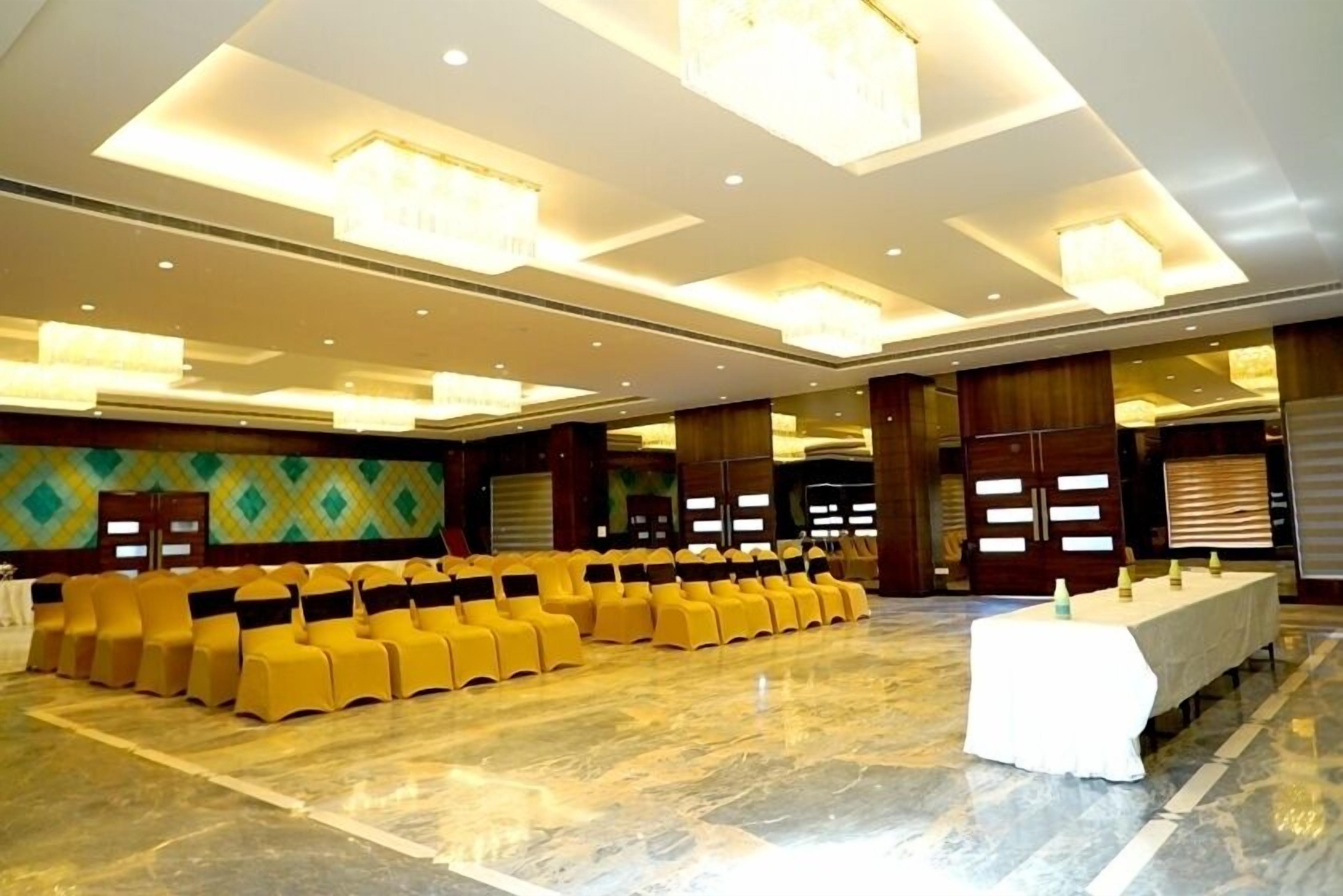 Uma sala de conferências com fileiras de cadeiras amarelas, uma mesa comprida com toalha branca e lustres no teto.