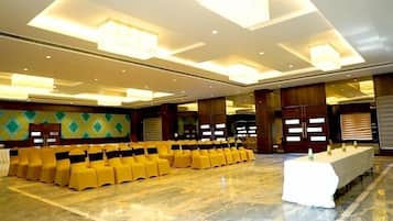 Uma sala de conferências com fileiras de cadeiras amarelas, uma mesa comprida com toalha branca e lustres no teto.