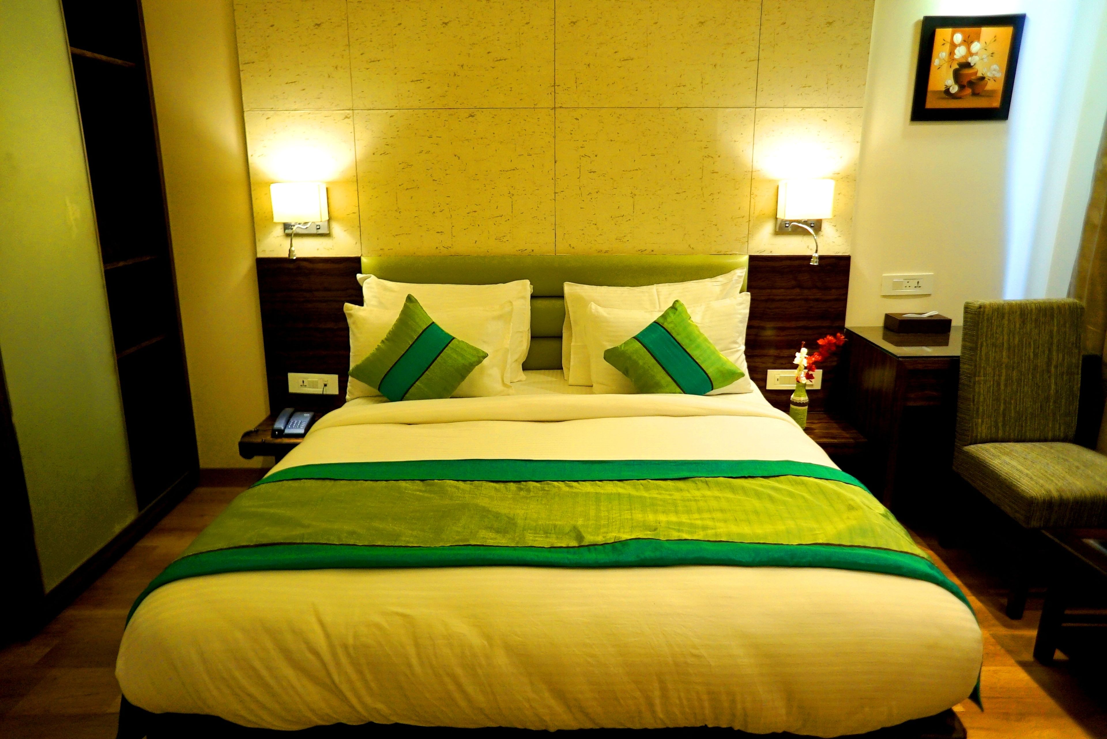Quarto de hotel com cama grande, roupa de cama verde e branca, duas luminárias de cabeceira, mesa de cabeceira com telefone, uma cadeira e uma pequena mesa com um vaso de flores.