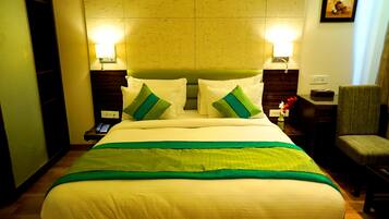 Quarto de hotel com cama grande, roupa de cama verde e branca, duas luminárias de cabeceira, mesa de cabeceira com telefone, uma cadeira e uma pequena mesa com um vaso de flores.