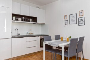 Uma cozinha moderna com armários brancos, pia de aço inoxidável e um espaço de refeições com mesa branca e cadeiras cinzas.