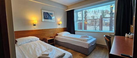 Quarto de hotel com duas camas, cabeceira de madeira, janela com vista para árvores e uma mesa com uma cadeira.