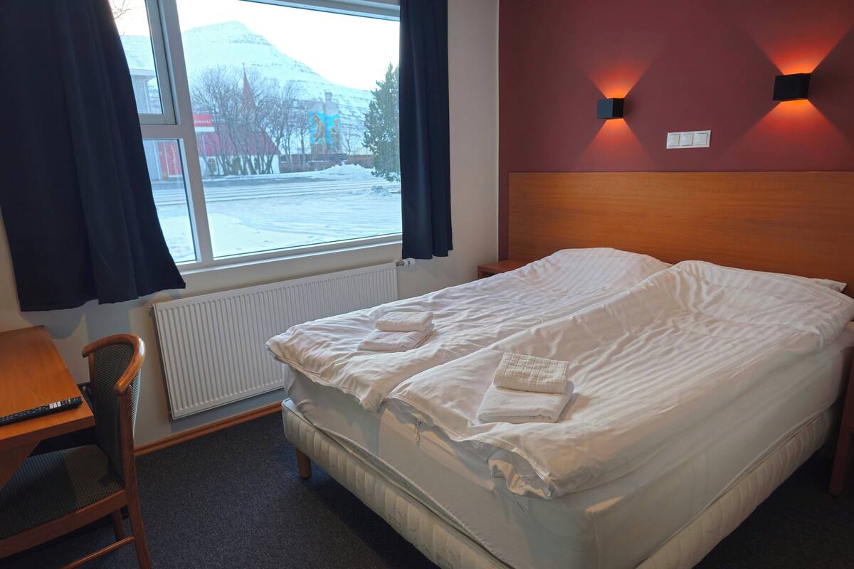Quarto de hotel com cama, cadeira, escrivaninha, janela e luminária fixada na parede.