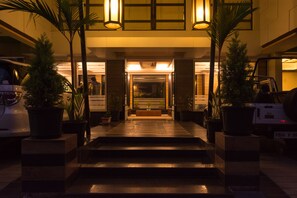 Entrada de hotel com uma escadaria que leva a uma área de recepção, ladeada por plantas em vasos e iluminada por luzes pendentes.