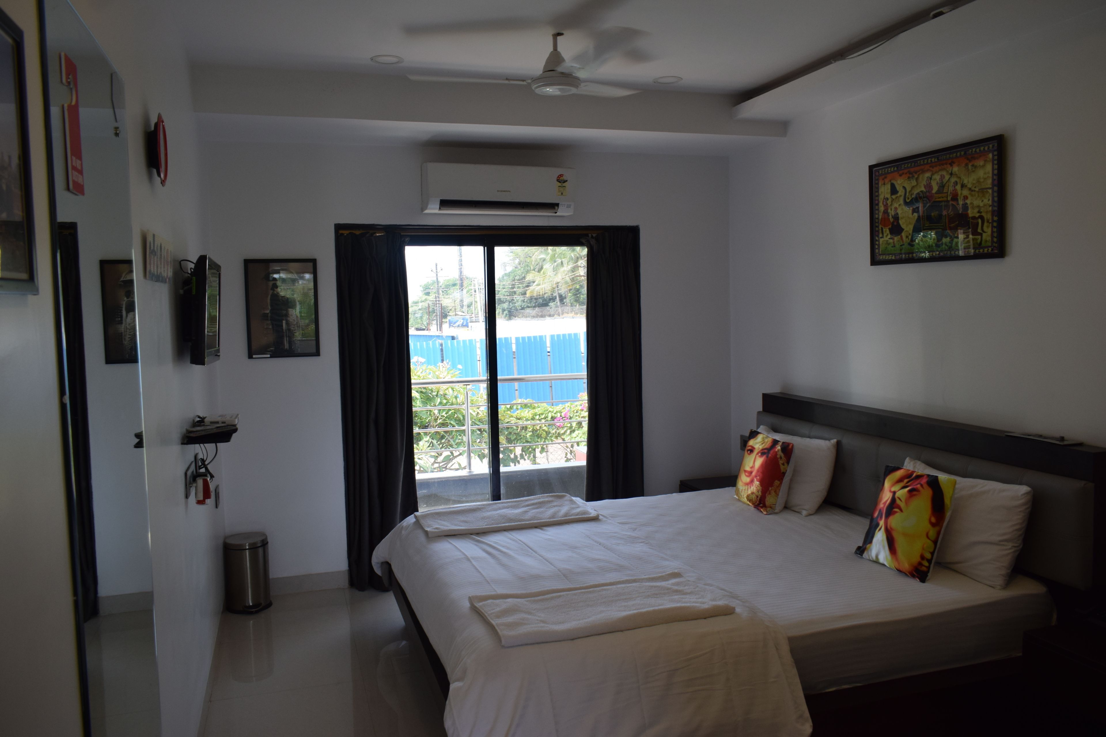 Quarto de hotel com cama, ventoinha no teto, ar condicionado, janela com cortinas e quadro pendurado na parede.