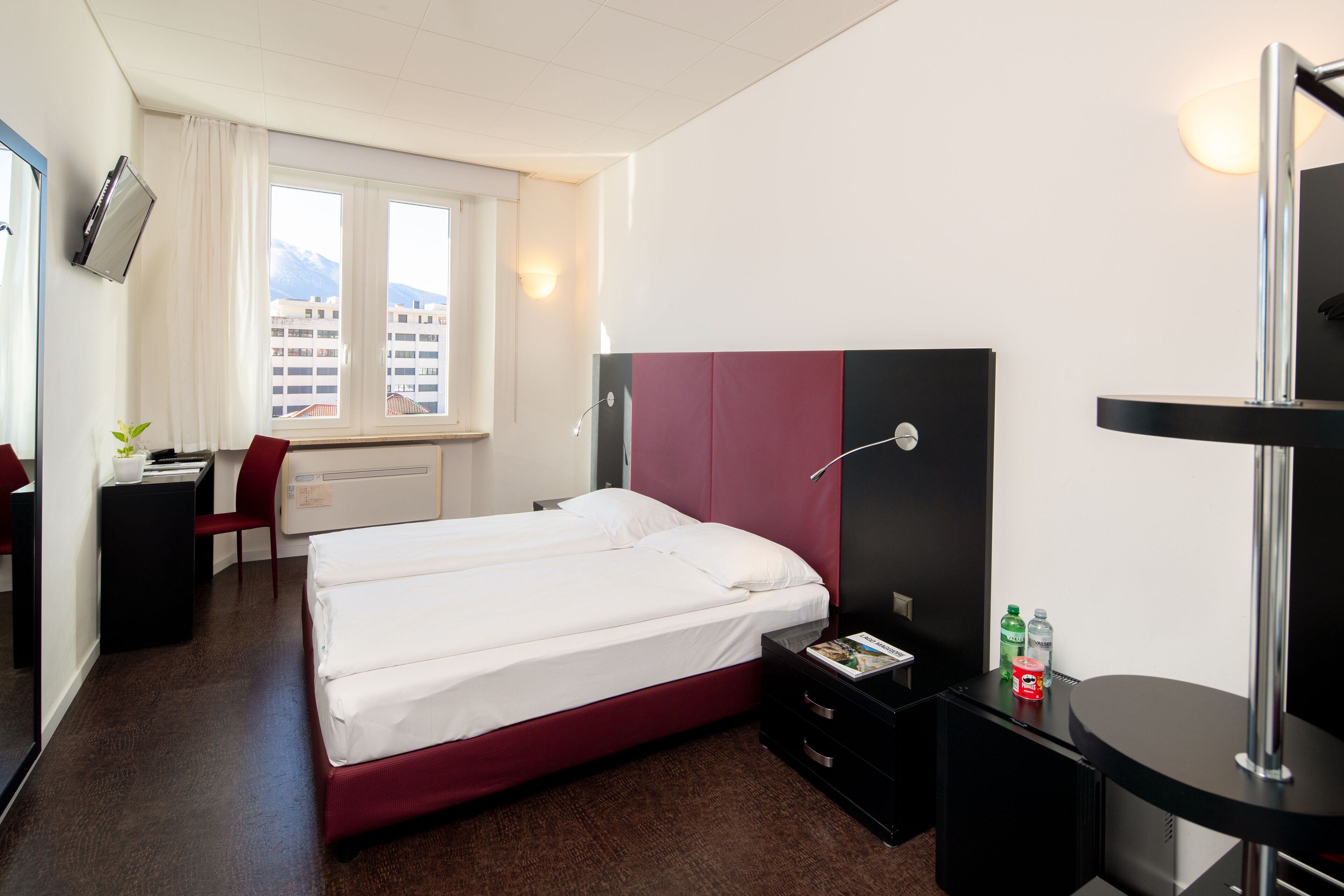 Quarto de hotel com cama, escrivaninha, cadeira, televisão e janela com vista para edifícios e montanhas.