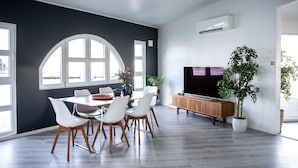 Uma sala de jantar moderna com uma grande janela, cadeiras brancas, uma mesa de jantar de madeira e uma TV sobre um móvel de madeira.