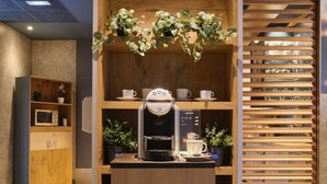 Uma máquina de café Nespresso em uma vitrine de madeira, com uma xícara e pires, cercada por prateleiras com plantas e um micro-ondas.