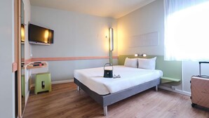 Quarto de hotel com cama, mesa de cabeceira, luminária, televisão e mala.