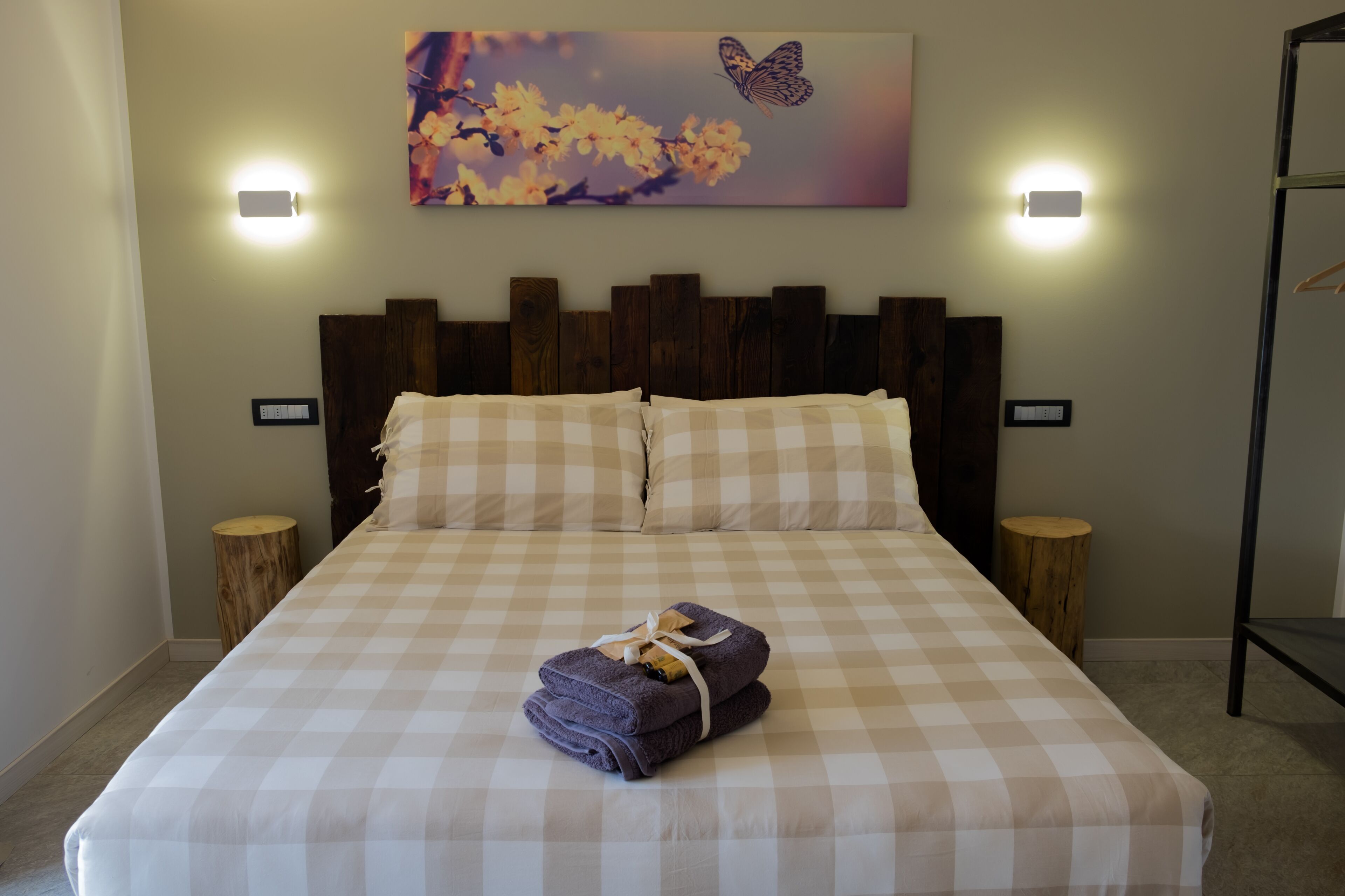 Uma cama bem arrumada com roupa de cama xadrez, duas mesas de cabeceira, luminária fixada na parede e um quadro de uma borboleta sobre a cama.