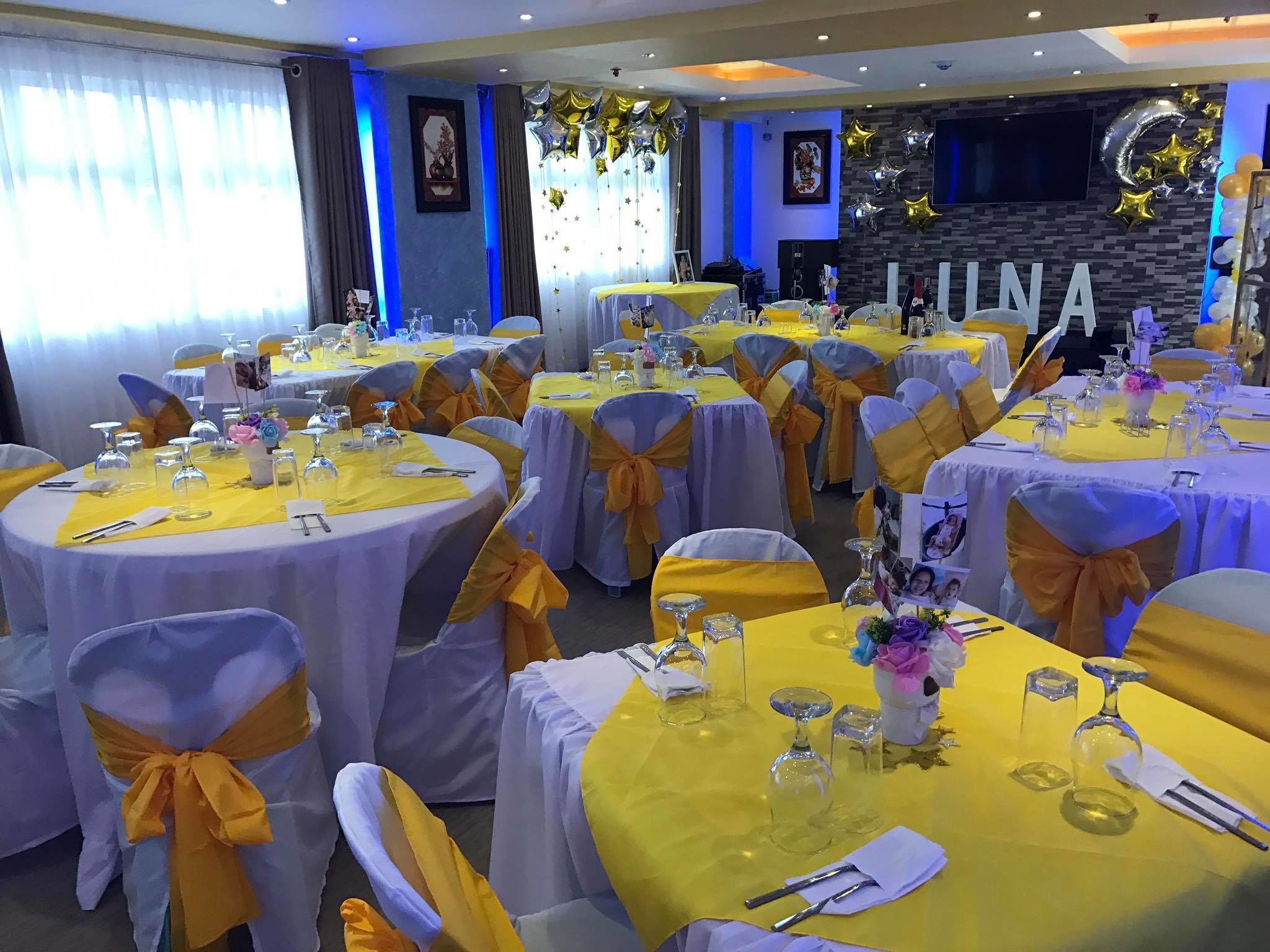 Salão de festas com mesas redondas, toalhas de mesa amarelas e brancas, e cadeiras com fitas amarelas.