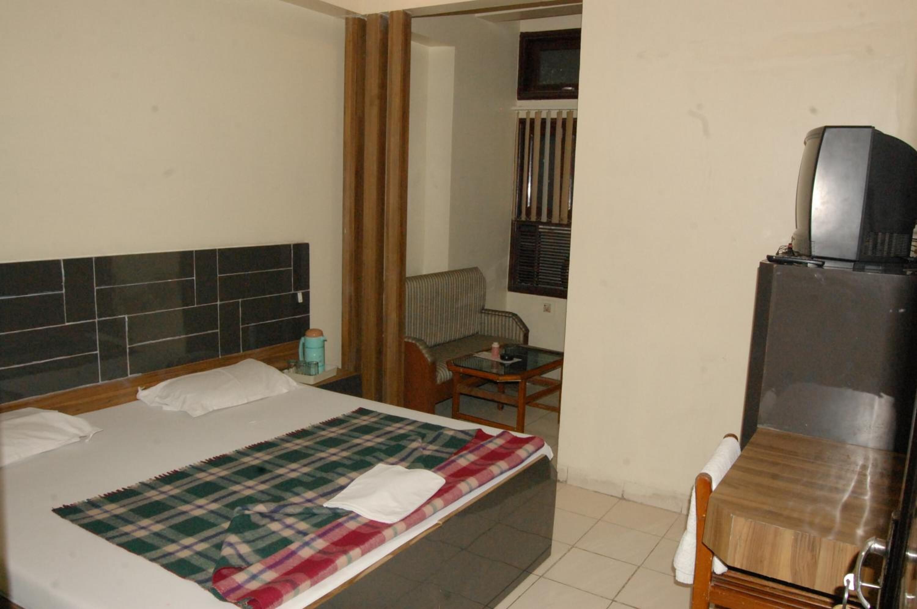 Quarto de hotel com cama, mesa pequena, cadeira, televisão e janela com persianas.