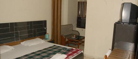 Quarto de hotel com cama, mesa pequena, cadeira, televisão e janela com persianas.